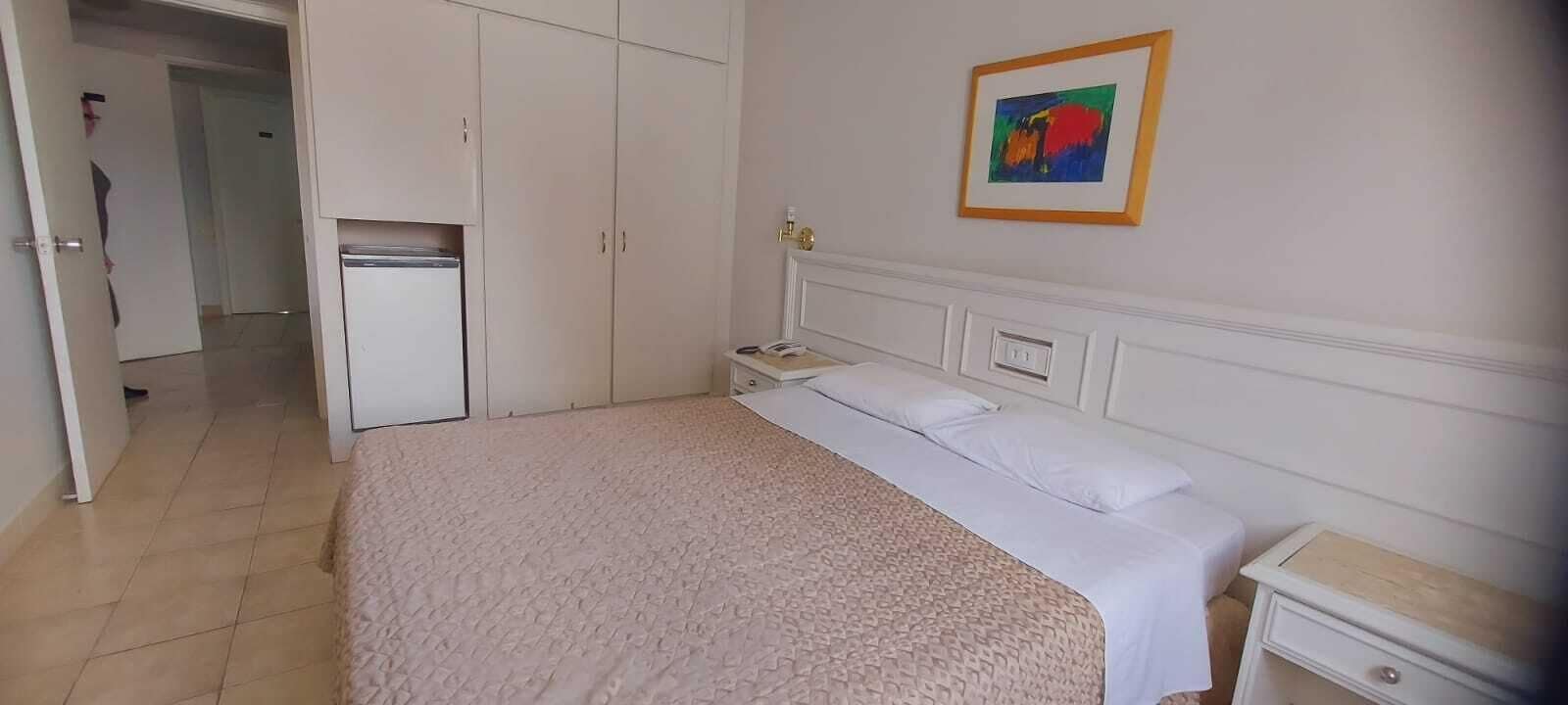 Quarto com cama, mesa de cabeceira, cômoda e porta que dá para outro espaço.