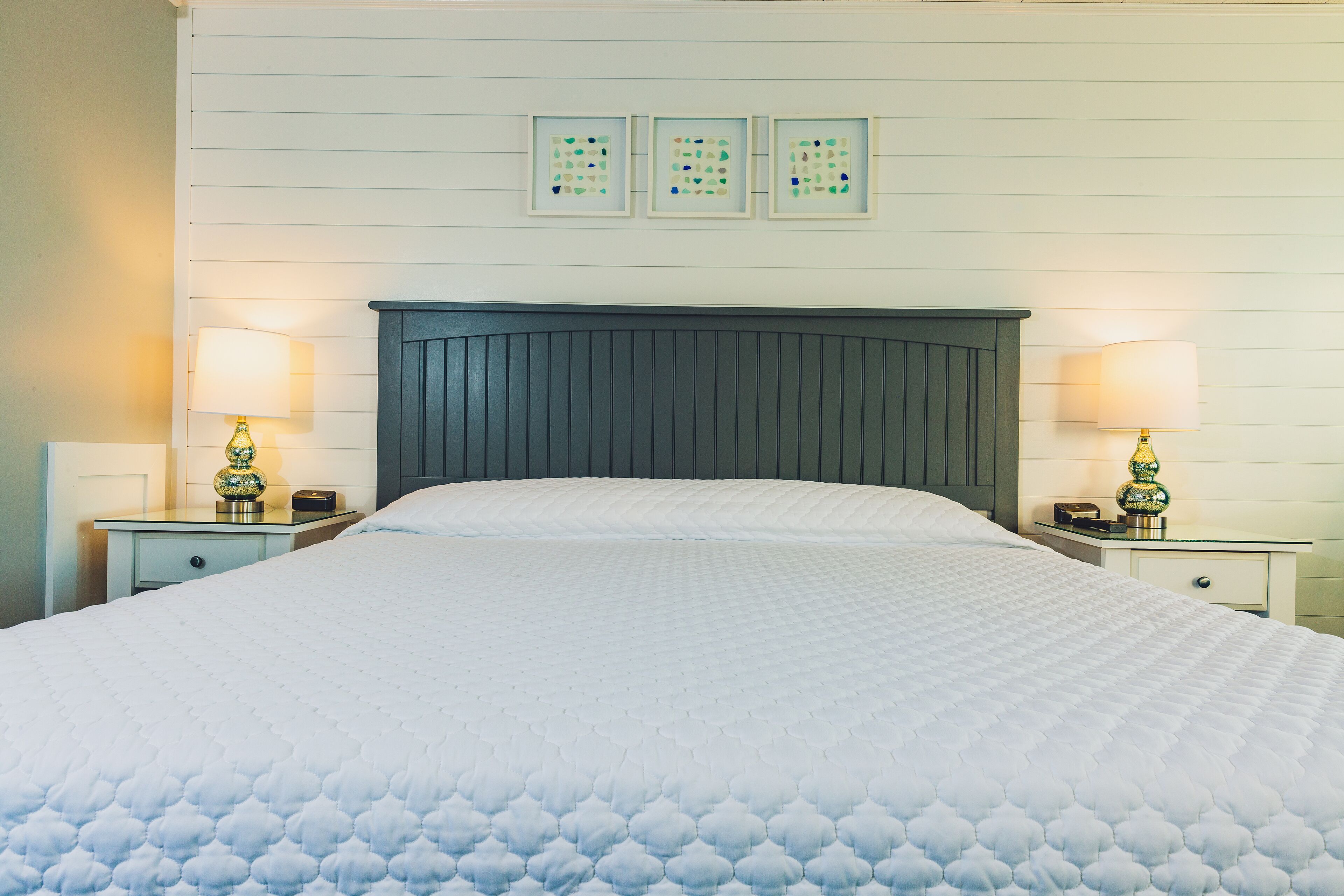 Cama bem arrumada com edredão branco acolchoado, duas mesas de cabeceira com abajures e três quadros na parede acima da cama.