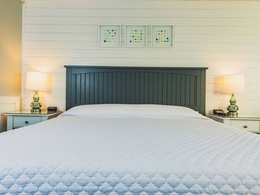 Cama bem arrumada com edredão branco acolchoado, duas mesas de cabeceira com abajures e três quadros na parede acima da cama.