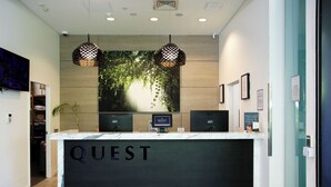 Uma receção com o nome "QUEST" em destaque, duas luzes pendentes, uma televisão e certificados emoldurados na parede.