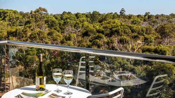 Mesa com garrafa e copos, rodeada por cadeiras brancas, com vista para uma paisagem arborizada.