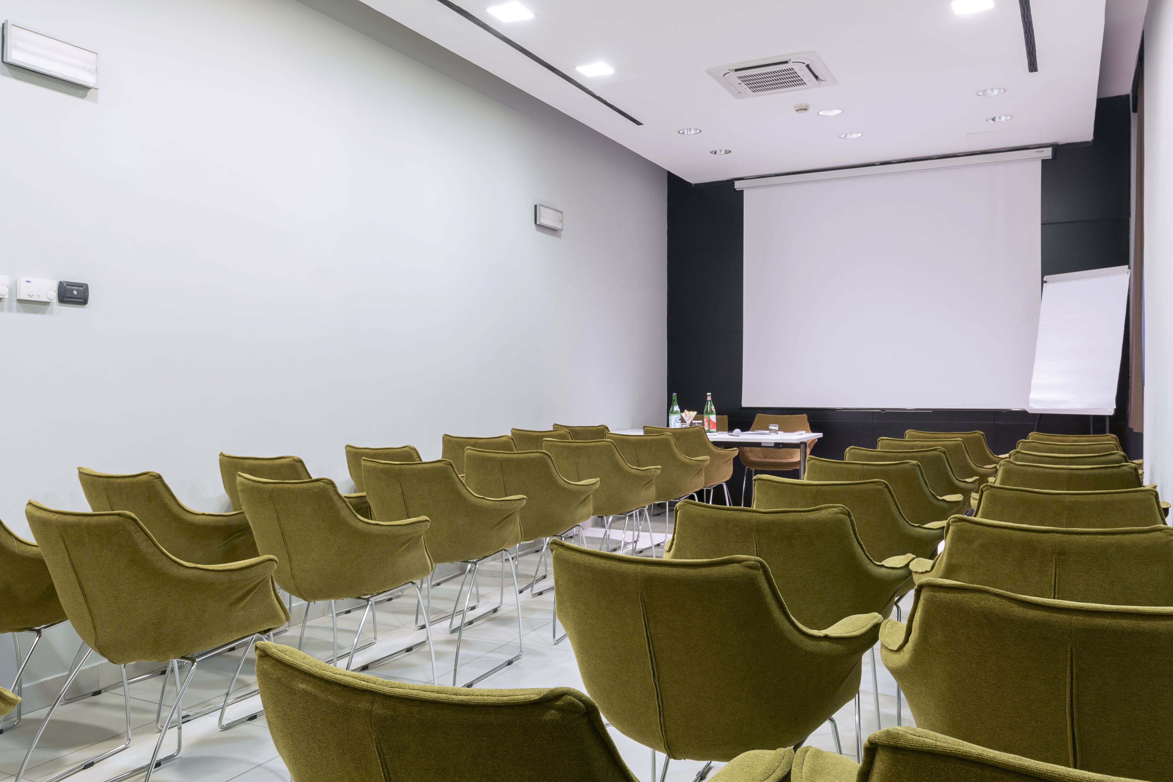Uma sala de conferências com fileiras de cadeiras verdes, uma tela de projetor e uma mesa para apresentações.