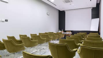 Uma sala de conferências com fileiras de cadeiras verdes, uma tela de projetor e uma mesa para apresentações.