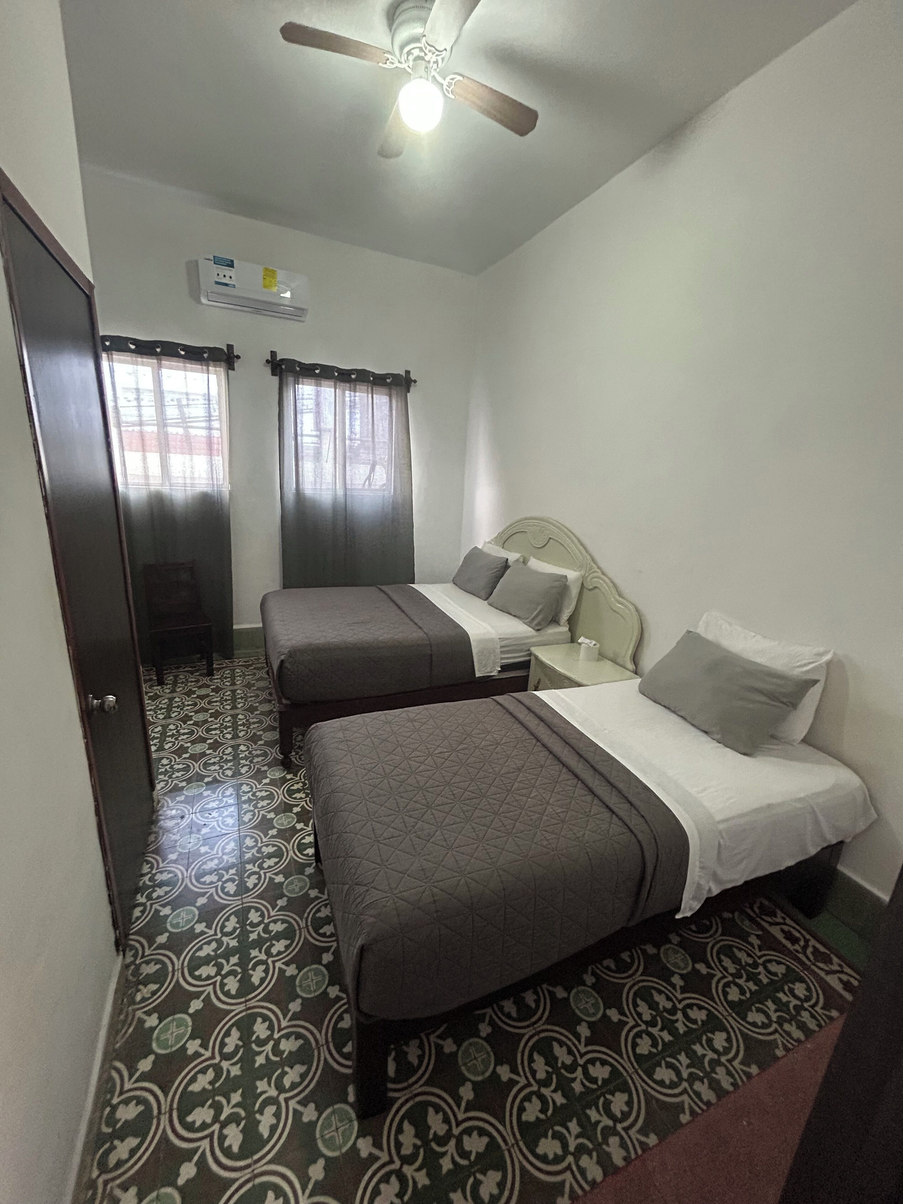 Quarto com duas camas, ventoinha no teto e piso de azulejos estampados.