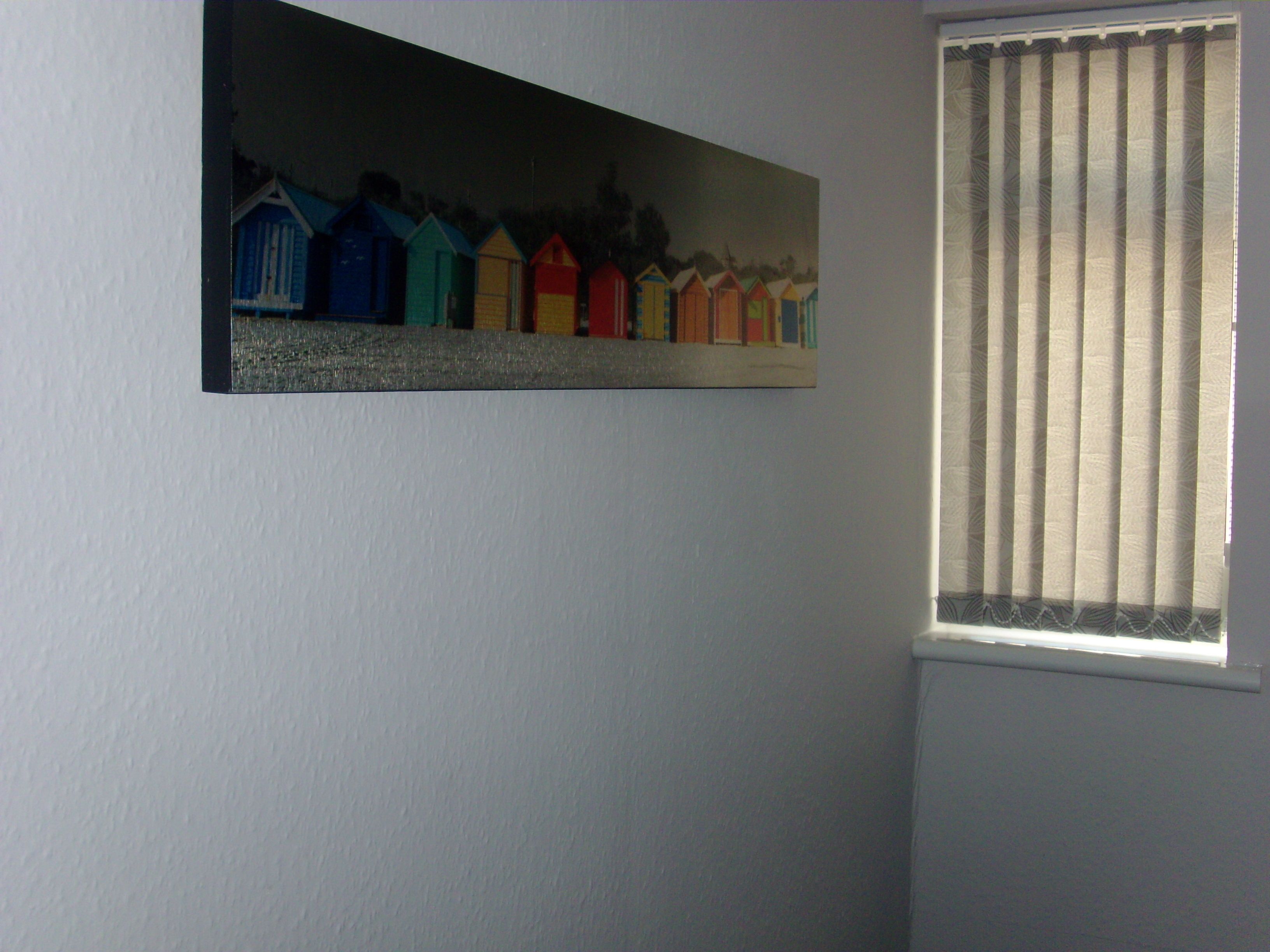 Um quarto com parede branca, janela com persianas verticais e uma pintura colorida de cabanas de praia.