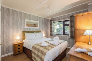 Quarto de hotel com cabeceira de madeira, cama grande com roupa de cama branca, mesa de cabeceira com abajur e janela com cortinas.