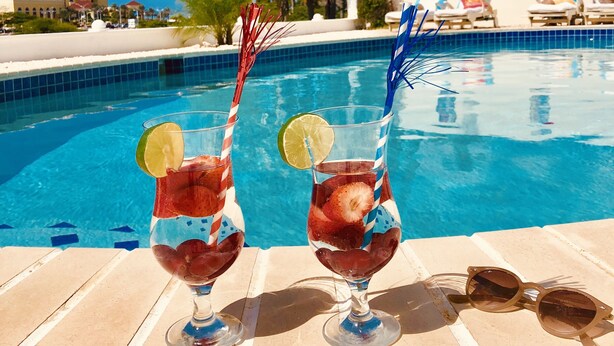 Dois cocktails com fatias de limão e canudos, perto de uma piscina com palmeiras e espreguiçadeiras ao fundo.