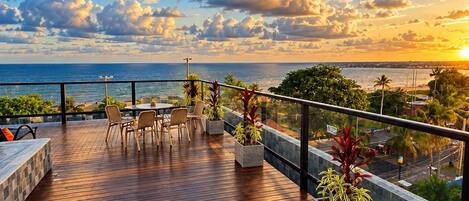 Um deck de madeira com mobiliário de exterior e plantas em vasos, com vista para um corpo d'água e a paisagem urbana.