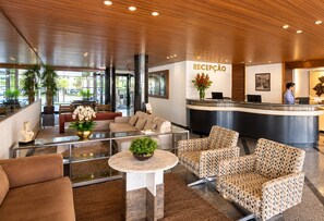 Área de recepção de hotel, com balcão de atendimento, espaço para sentar e recepcionista.