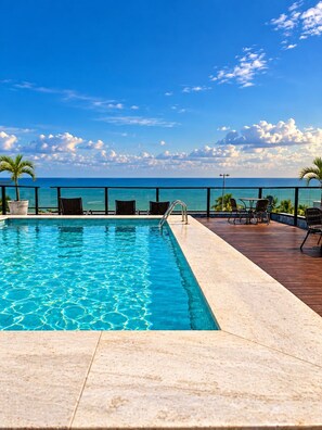 Piscina com vista para o oceano, rodeada por espreguiçadeiras e um deck de madeira.