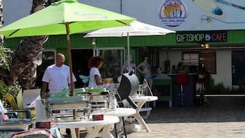 Um café ao ar livre com mesas e cadeiras, um guarda-sol verde e um letreiro de loja de presentes.