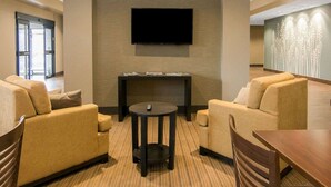 Um lobby de hotel com uma televisão de tela plana na parede, uma mesa pequena com um controle remoto e dois sofás com uma mesa de madeira e cadeiras em primeiro plano.