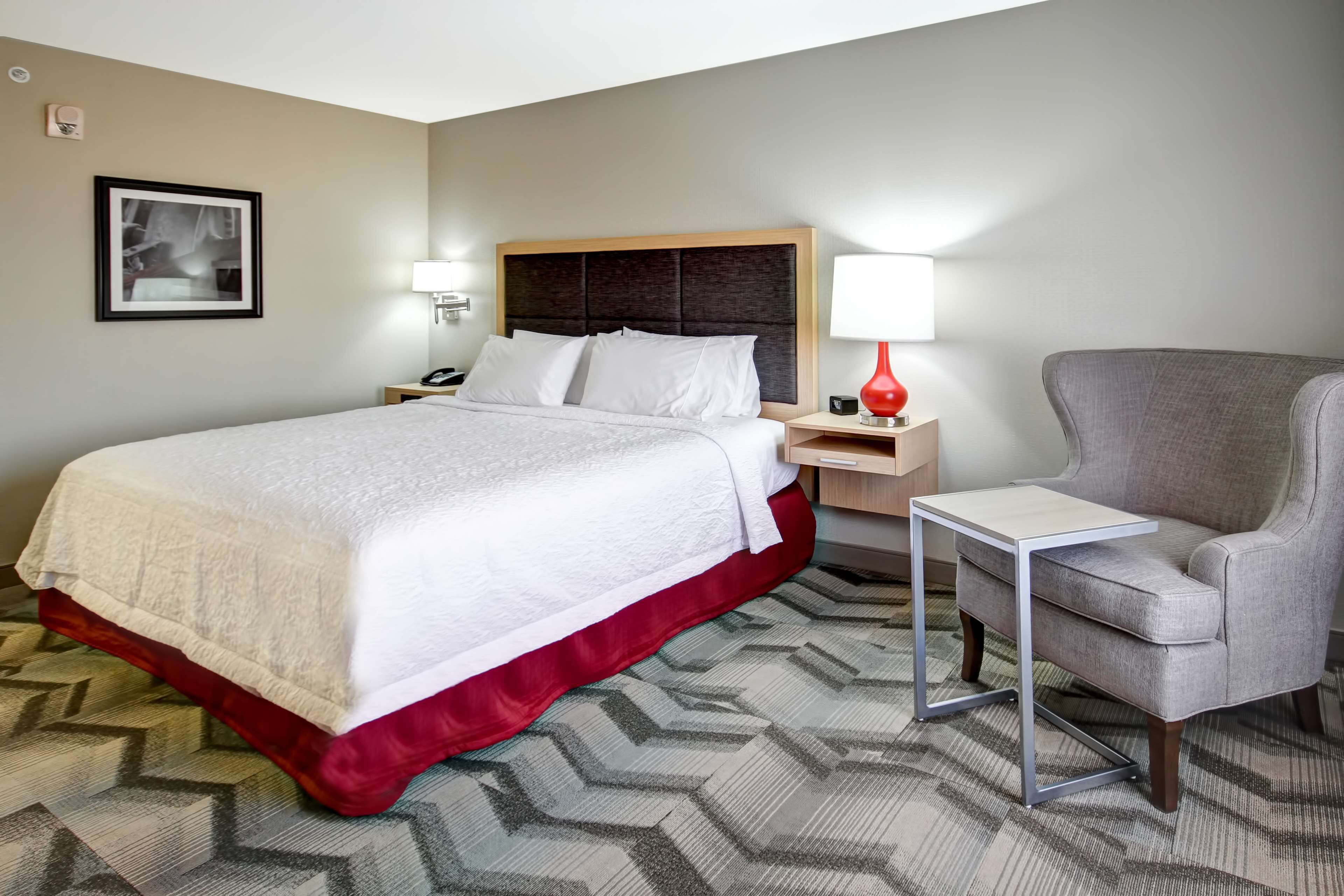 Quarto de hotel com uma cama grande, uma cadeira, uma mesinha e uma mesa de cabeceira com abajur.