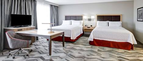 Quarto de hotel com duas camas, uma mesa de trabalho com televisão, uma cadeira e abajur.