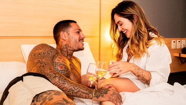 Um casal sentado em uma cama em quarto de hotel, segurando taças de vinho.