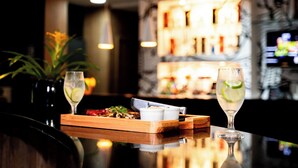 Uma área de bar com uma mesa de madeira, dois copos de uma bebida cítrica, um pequeno prato com comida e um vaso de planta.