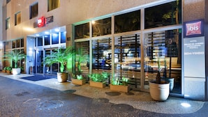 Entrada de hotel com grandes portas de vidro, plantas em vasos e placa indicando ibis hotel.