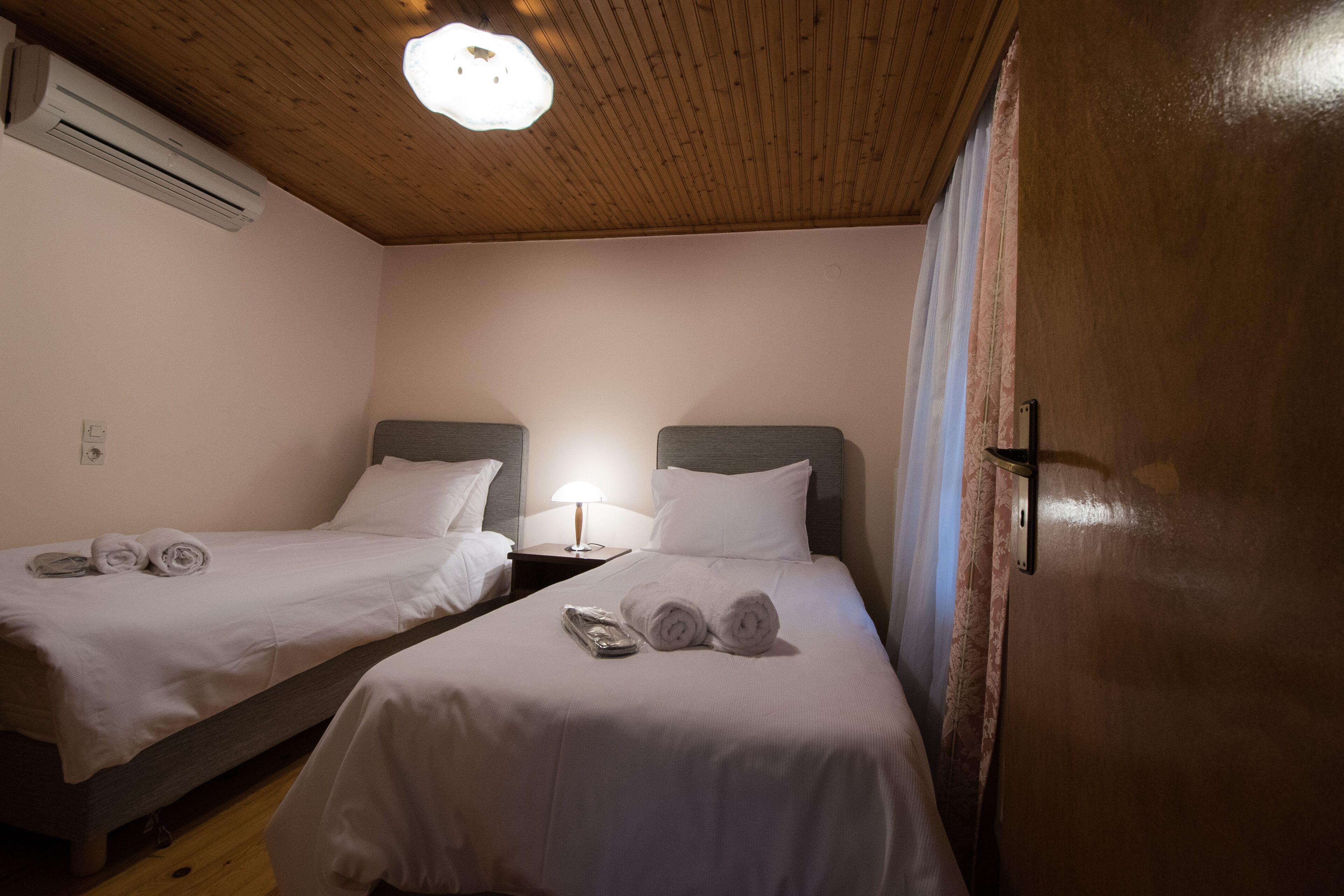 Quarto de hotel com duas camas, cada uma com roupa de cama branca e toalhas dobradas. Há uma mesa de cabeceira com uma luminária entre as camas, e um aparelho de ar condicionado fixado na parede.