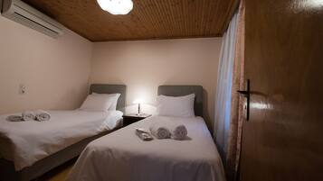 Quarto de hotel com duas camas, cada uma com roupa de cama branca e toalhas dobradas. Há uma mesa de cabeceira com uma luminária entre as camas, e um aparelho de ar condicionado fixado na parede.