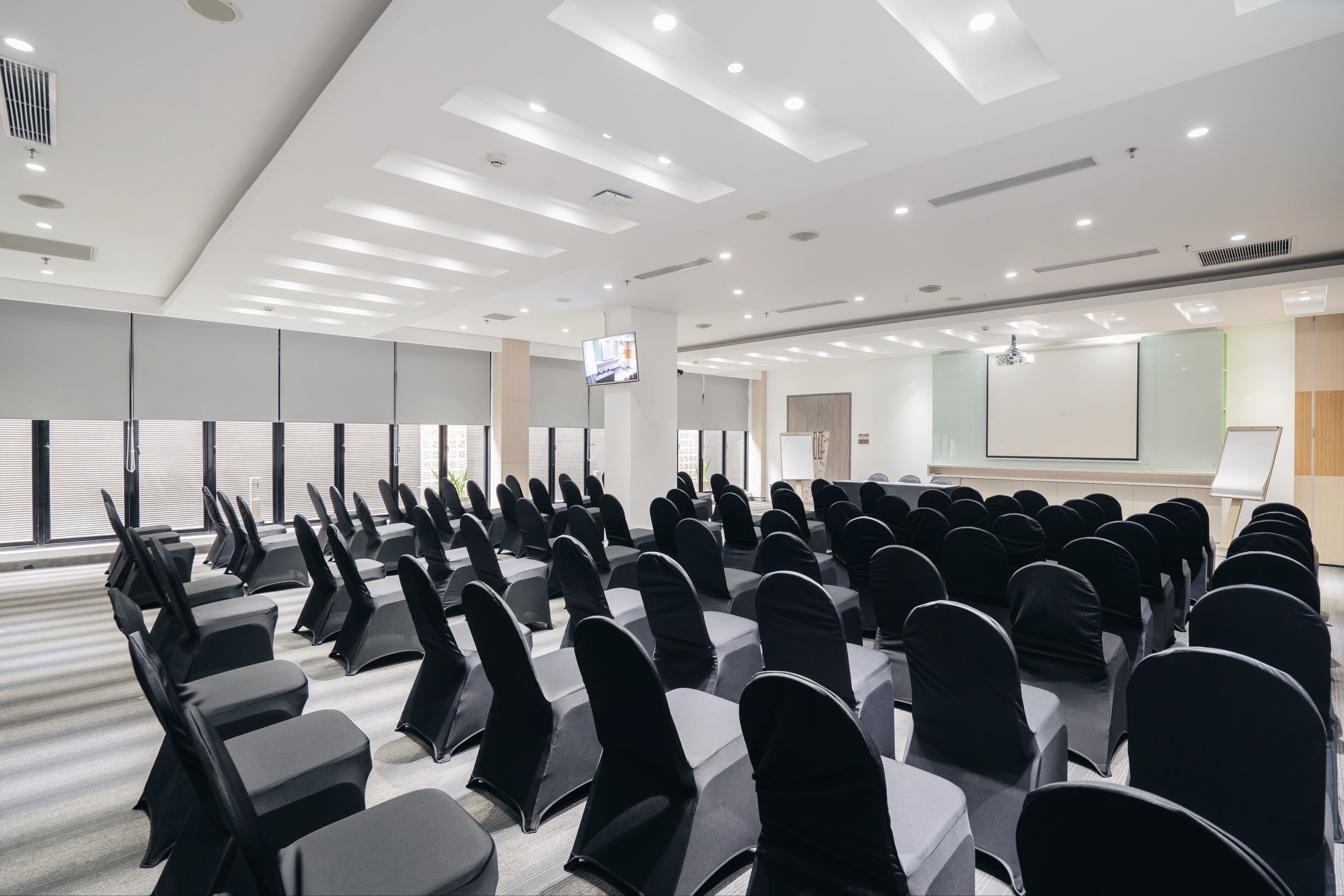 Sala de conferências com fileiras de cadeiras, uma tela de projeção e uma janela grande.