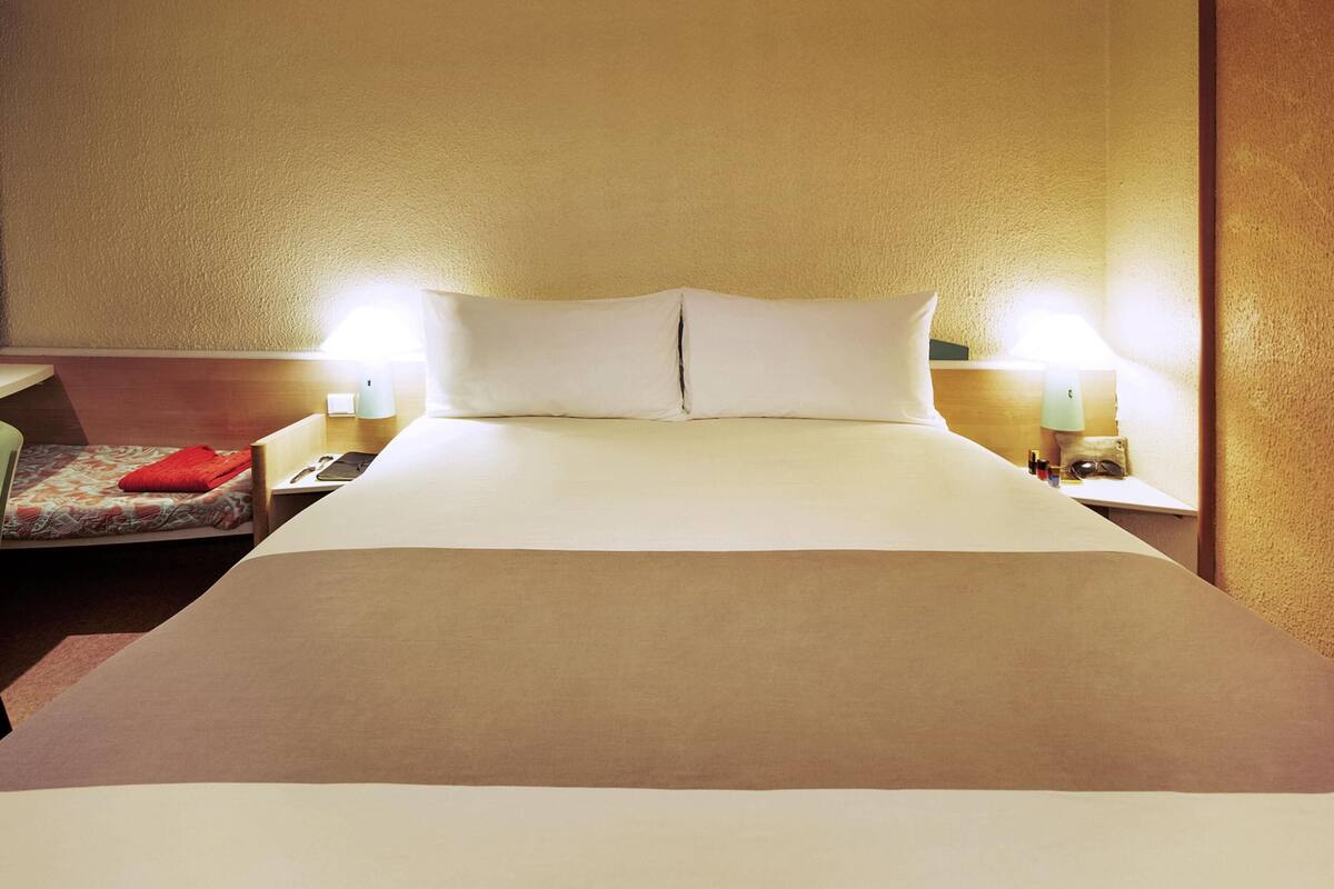 Quarto de hotel com uma cama grande, luminárias de cabeceira, uma mesinha e uma cadeira.