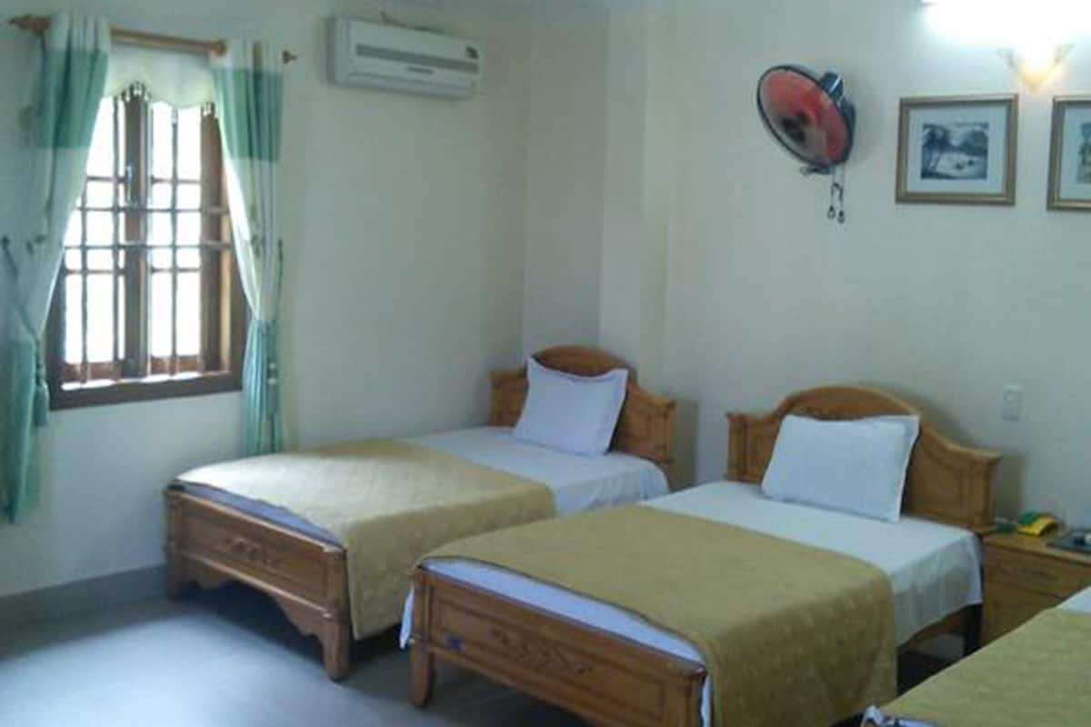 Quarto com duas camas de madeira, janela com cortinas, ar condicionado e ventoinha na parede.