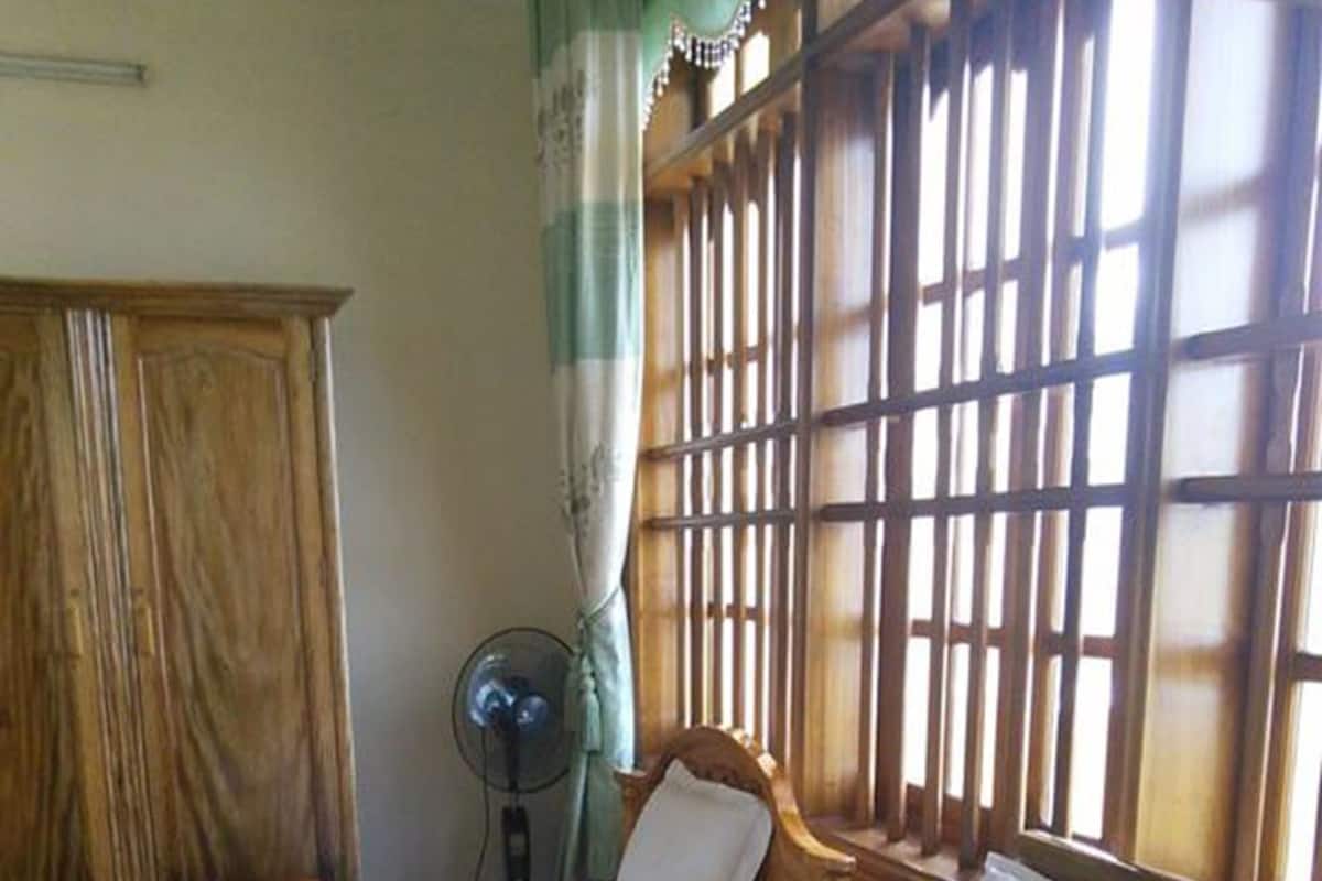 Quarto de hotel com duas camas, um guarda-roupa de madeira, um ventilador e uma janela com persianas verticais.