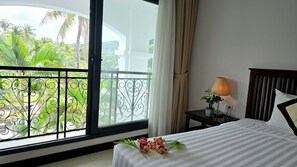 Quarto de hotel com uma janela grande, cama com roupa de cama listrada, mesa de cabeceira com abajur e vista para palmeiras e vegetação lá fora.