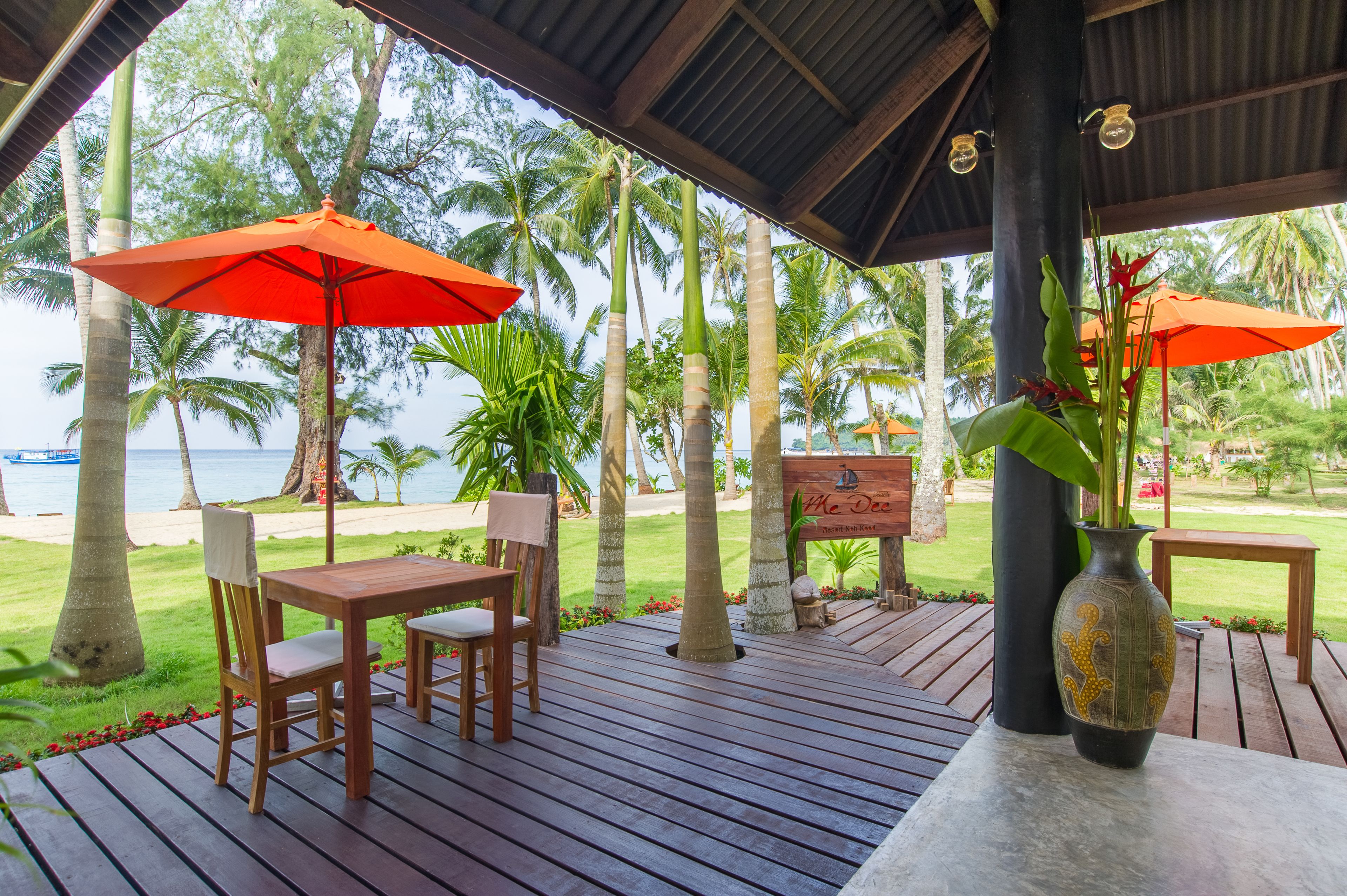 Um deck de madeira com mesa e cadeiras, guarda-sóis vermelhos e vista para a praia e palmeiras.
