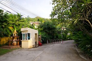 Uma entrada com portão e um pequeno quiosque, rodeada por vegetação exuberante e um edifício residencial ao fundo.