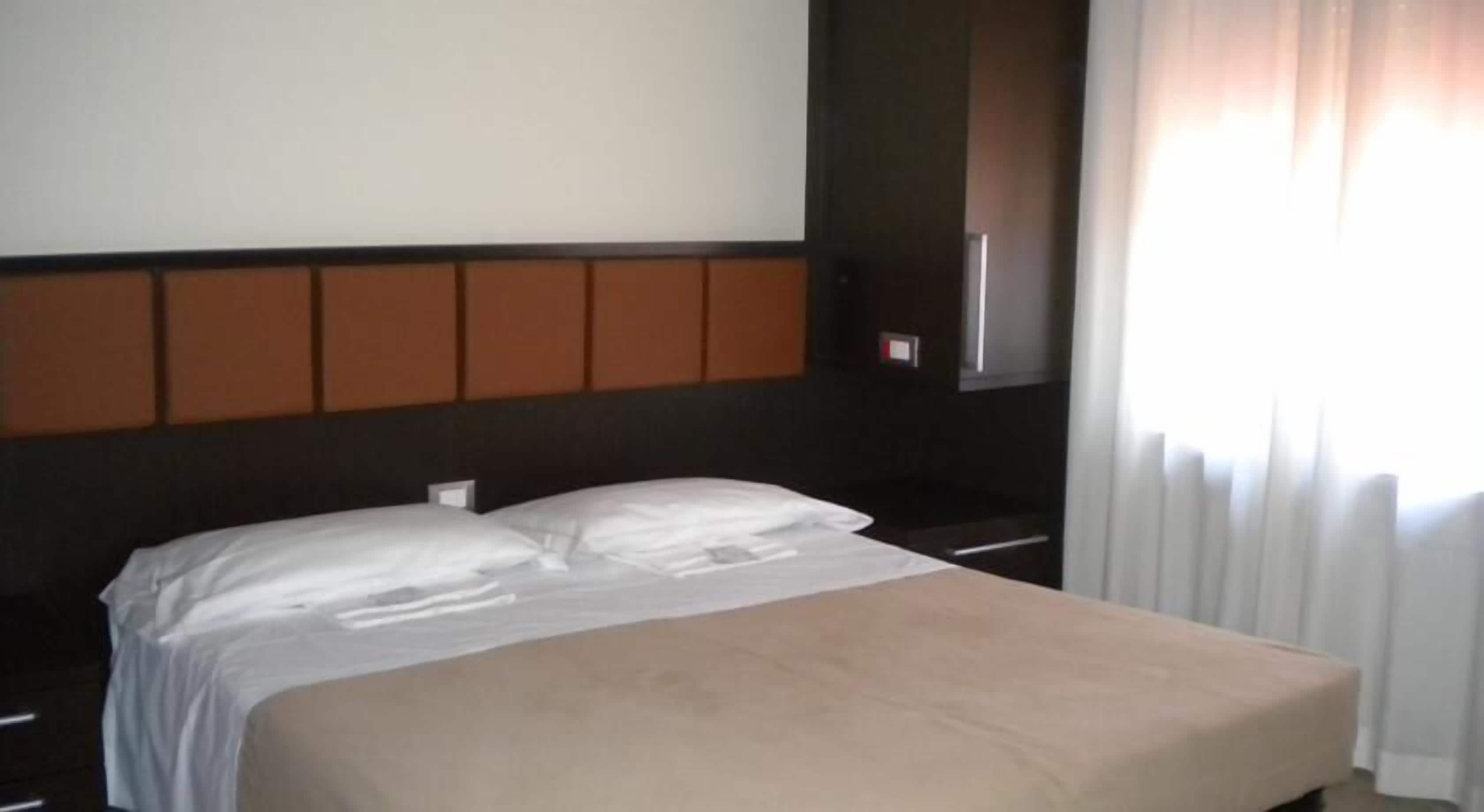Quarto de hotel com cama, cabeceira, mesa de cabeceira e janela com cortinas.