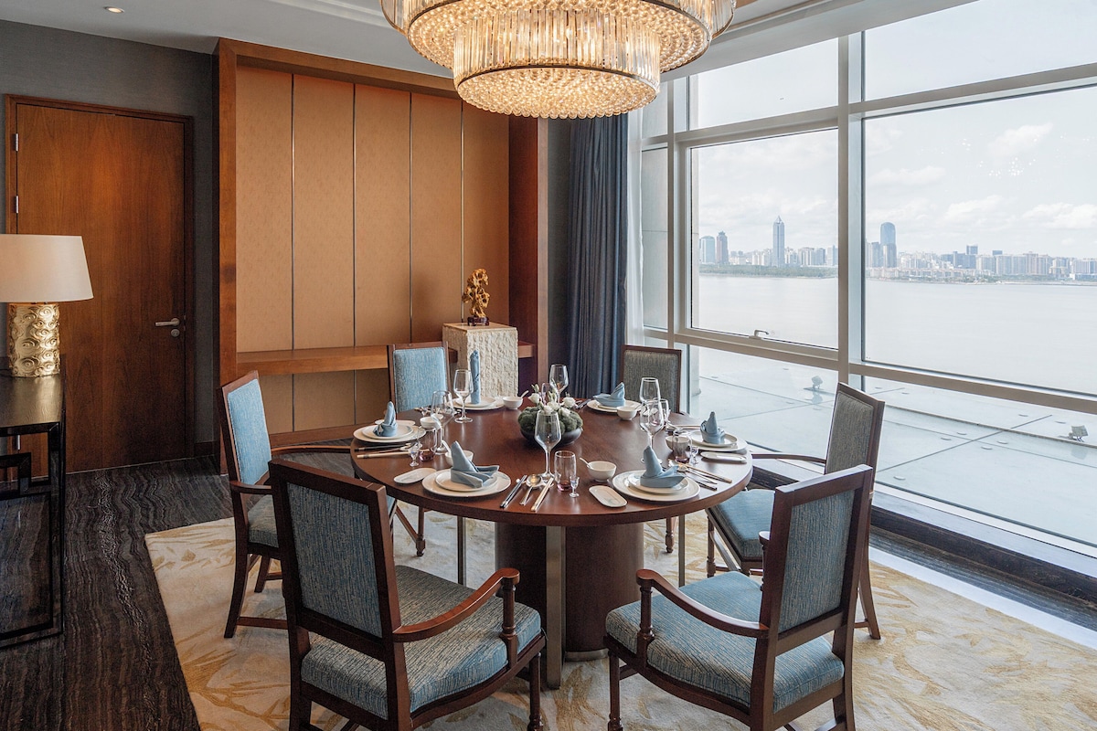 Uma sala de jantar com uma mesa redonda posta para seis, grandes janelas com vista para a cidade, um candelabro e uma porta de madeira.