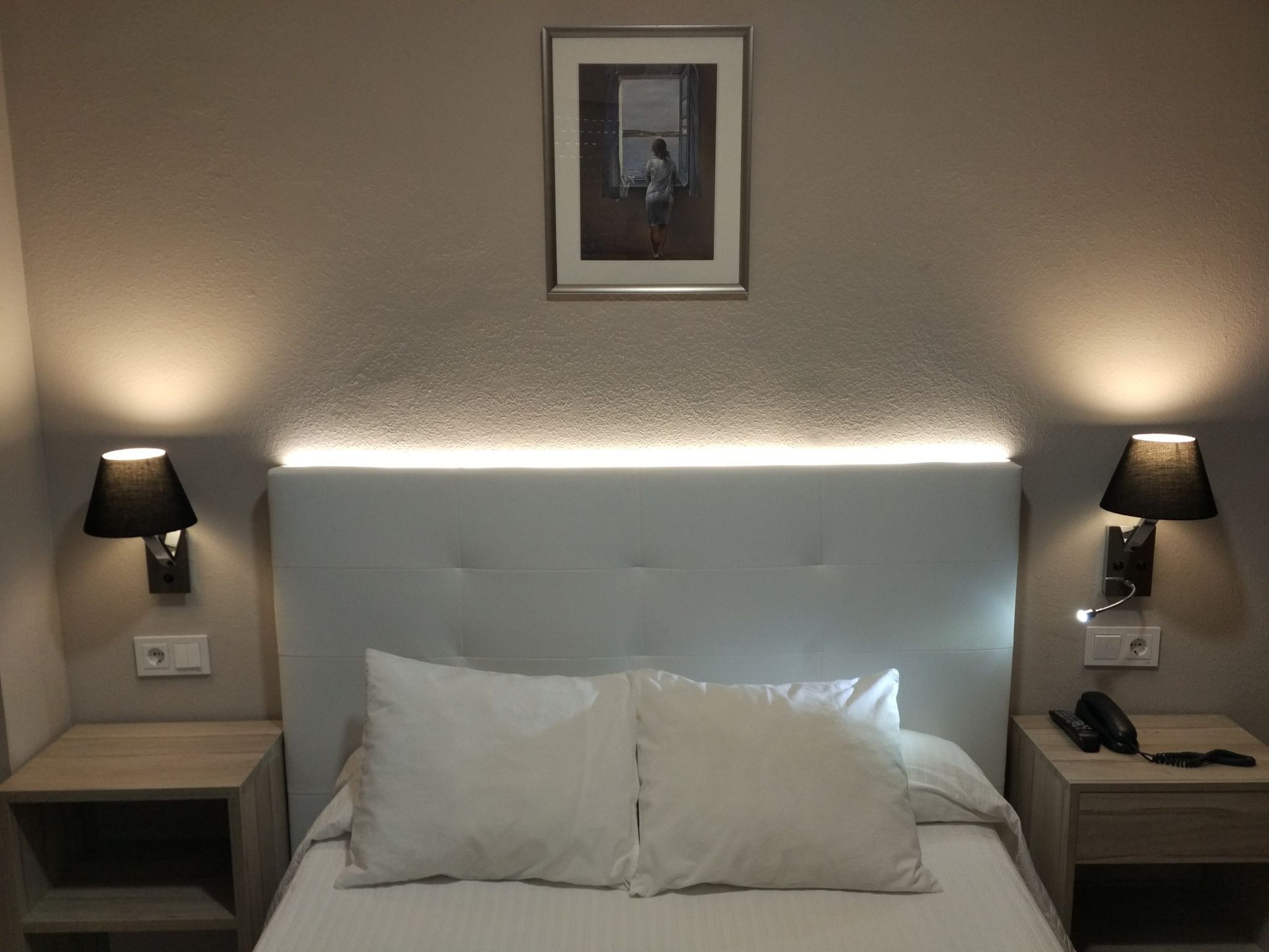 Quarto de hotel com uma cama, duas luminárias de cabeceira, um quadro na parede e uma mesa de cabeceira com telefone e controle remoto.