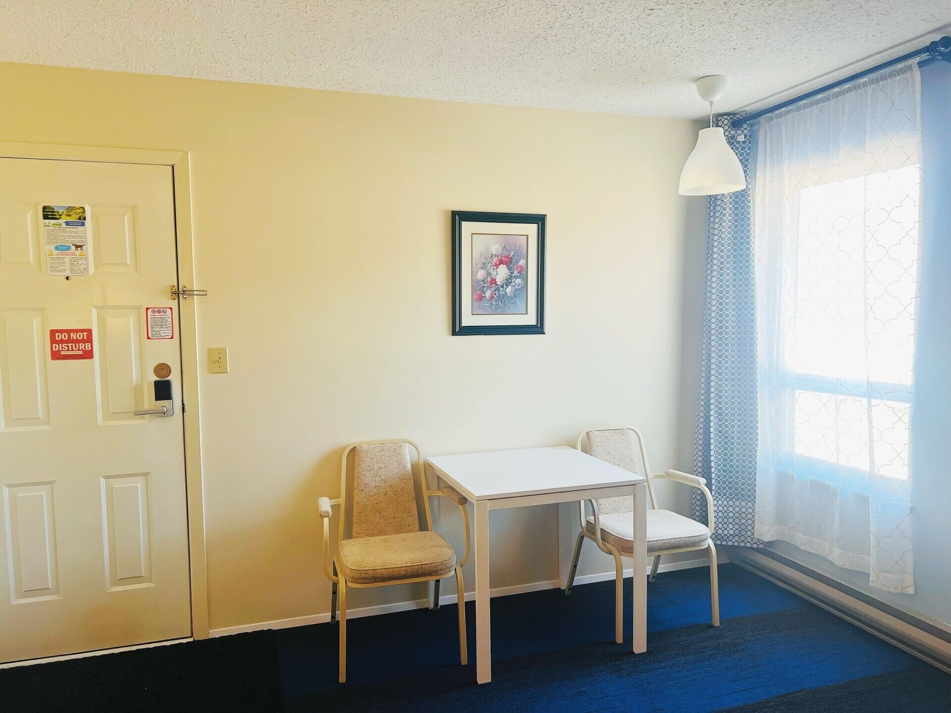 Um quarto com uma porta, duas cadeiras, uma mesa, um quadro emoldurado e uma janela com cortinas.