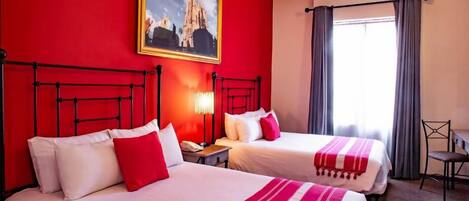 Quarto de hotel com duas camas, paredes vermelhas, um quadro de uma catedral e uma janela com cortinas.