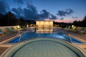 Uma piscina com deck revestido, rodeada por espreguiçadeiras verdes e uma área de estar coberta com mesas e cadeiras.