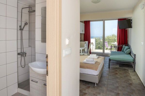 Casa de banho moderna com chuveiro, pia e espaço para guardar objetos. Um quarto adjacente possui uma cama, uma varanda com televisão e vista para o exterior.