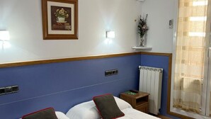 Quarto de hotel com duas camas, uma pequena mesa de cabeceira, um radiador, um quadro na parede e uma janela com cortinas.