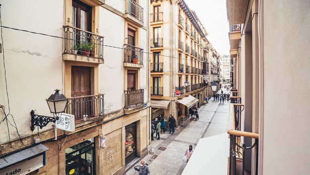 Uma rua estreita com edifícios em ambos os lados, com varandas adornadas com plantas, uma vitrine visível e um passeio para pedestres.