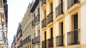 Uma rua estreita para pedestres, flanqueada por edifícios altos, com varandas e grades pretas, e plantas em vasos.