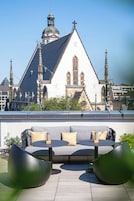 Um terraço no telhado com área de estar moderna, vista para uma igreja histórica com cúpula grande e torres, e céu azul límpido.