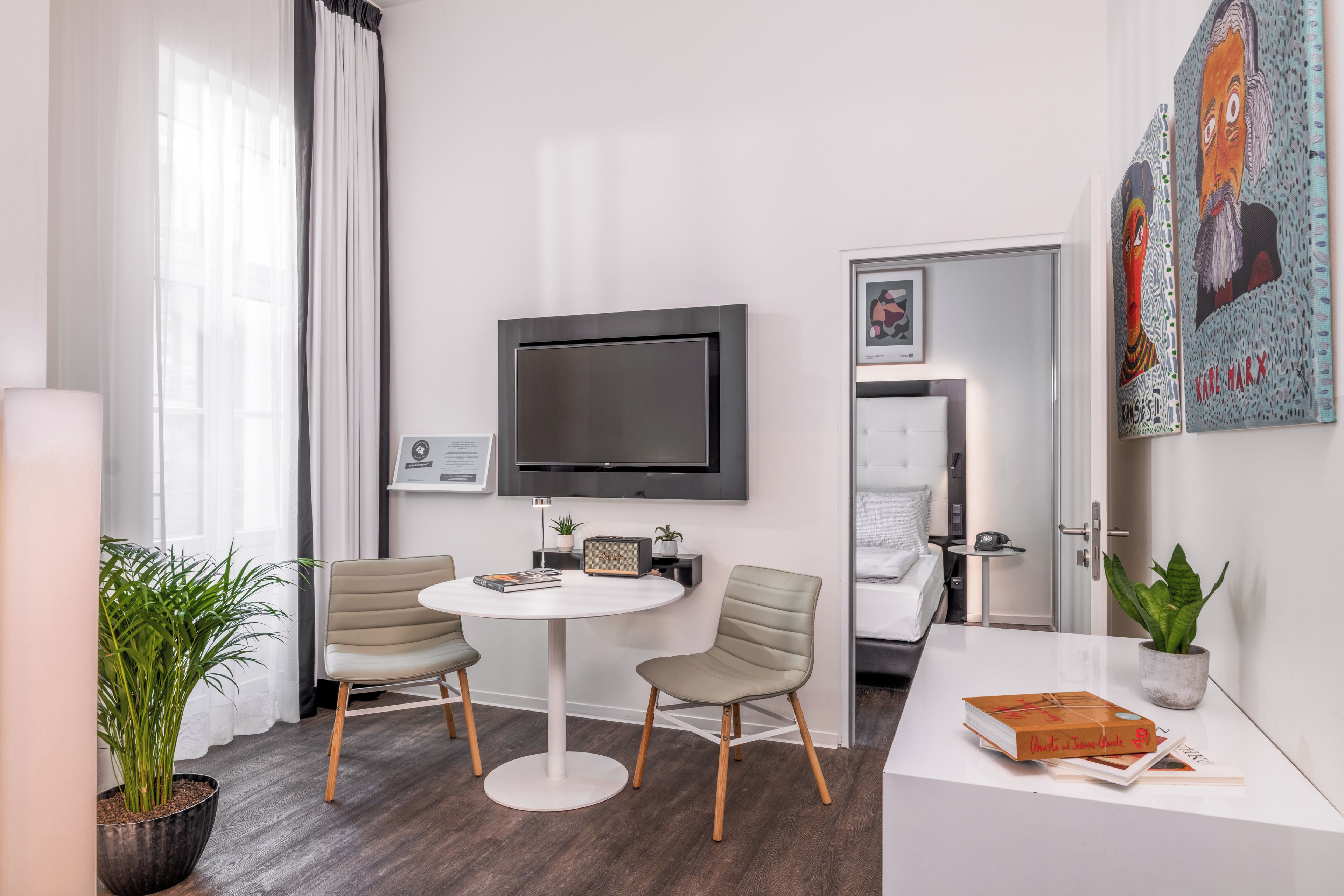 Quarto de hotel moderno com televisão de tela plana na parede, uma mesa de jantar redonda com duas cadeiras, uma planta em vaso e uma estante com livros.