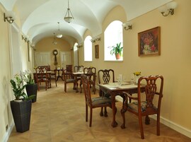 Uma área de refeições com cadeiras e mesas de madeira, um candelabro e uma janela grande com uma planta.