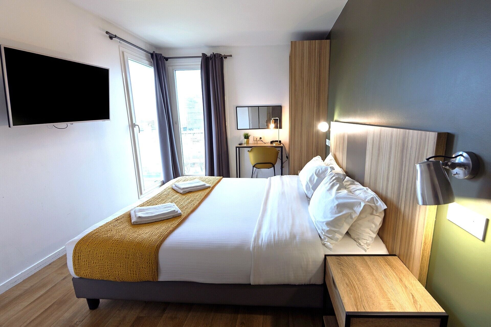 Quarto de hotel com cama, mesa de cabeceira, luminária, televisão e escrivaninha com cadeira.