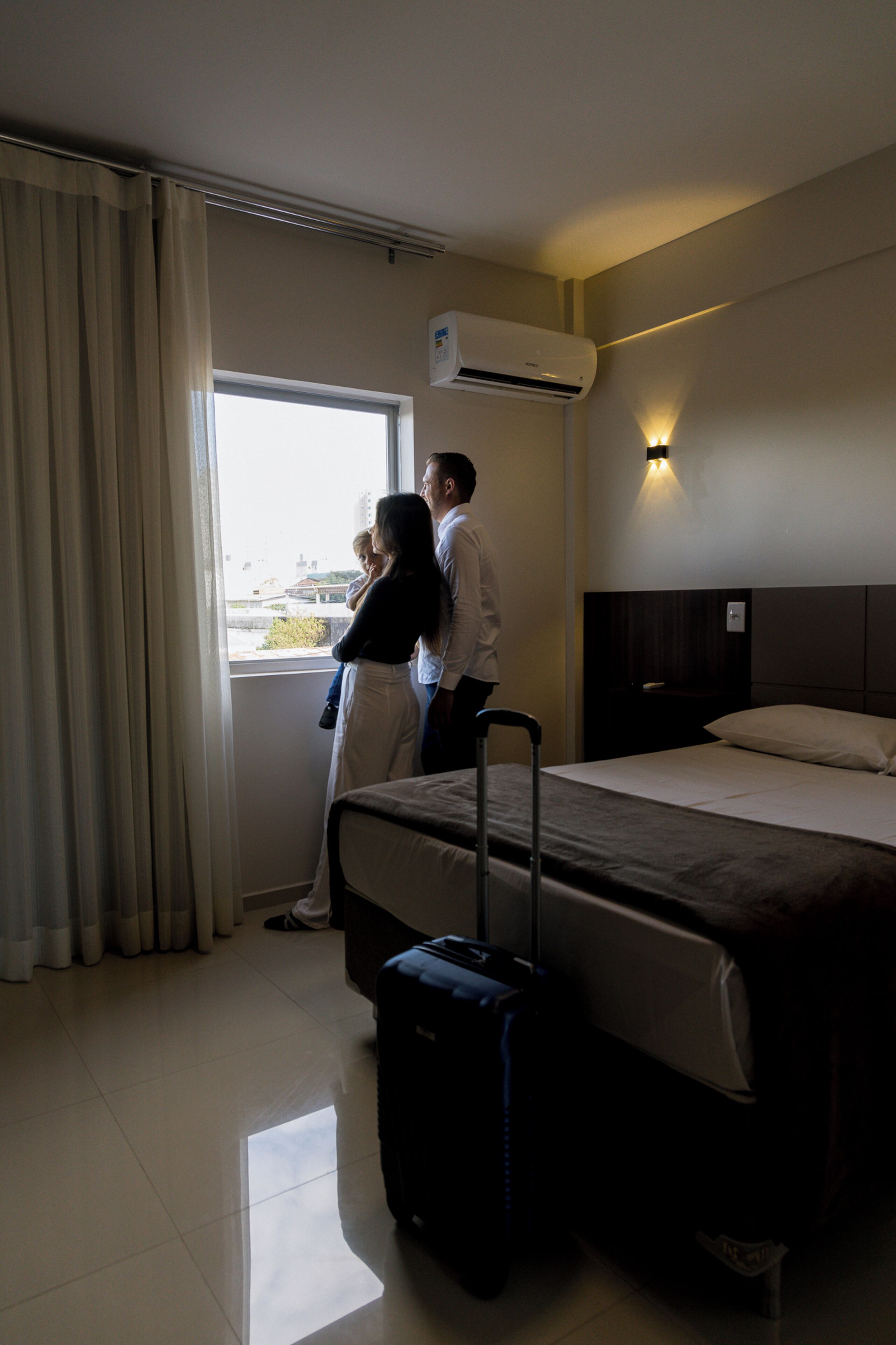Quarto de hotel com uma cama, uma mala e uma janela com cortinas.