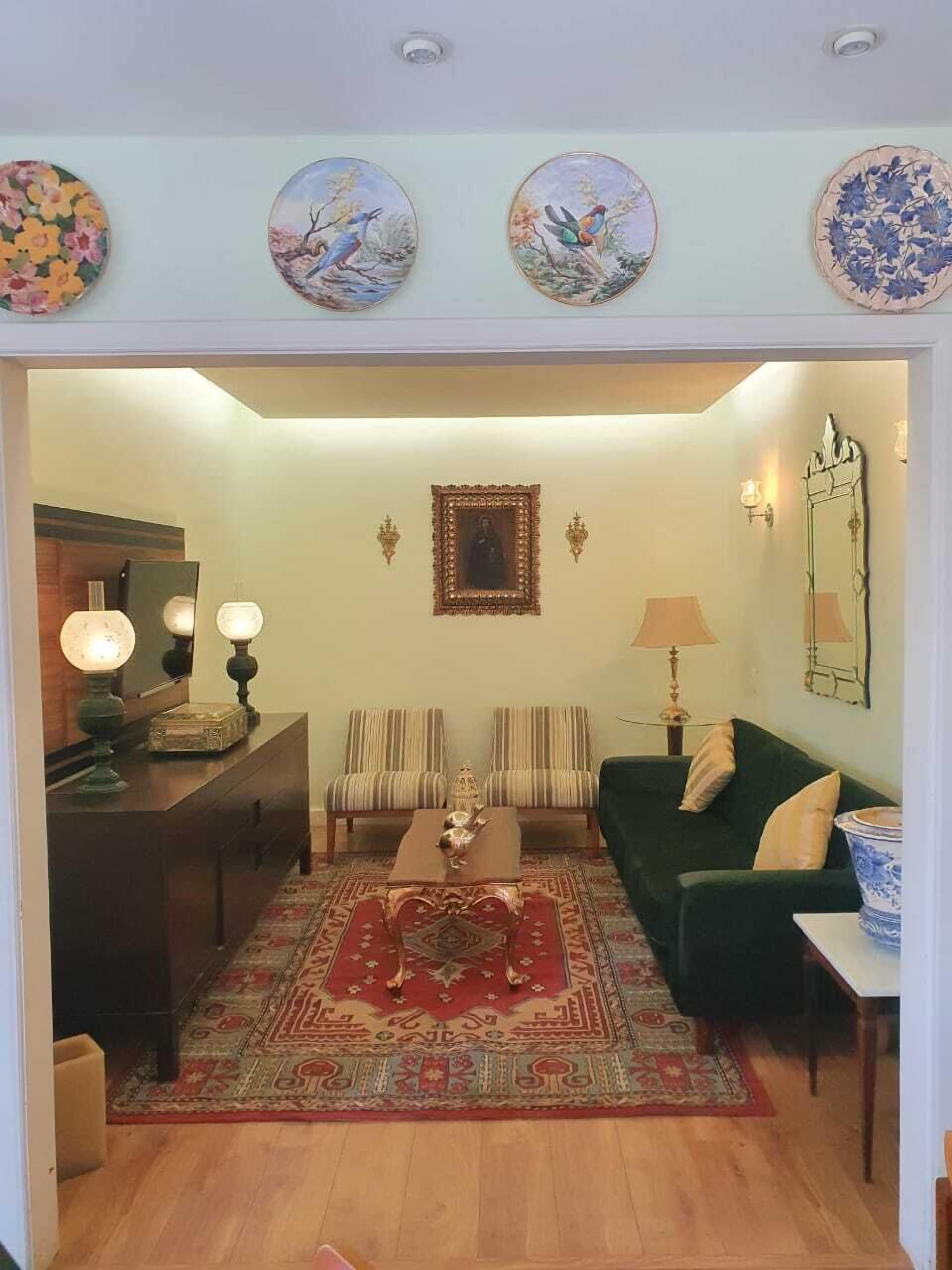 Uma sala de estar com um sofá verde, poltronas listradas, uma mesa de centro e um tapete estampado. Há luminárias fixadas na parede e um quadro emoldurado sobre o sofá.