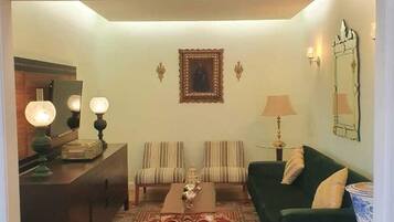 Uma sala de estar com um sofá verde, poltronas listradas, uma mesa de centro e um tapete estampado. Há luminárias fixadas na parede e um quadro emoldurado sobre o sofá.