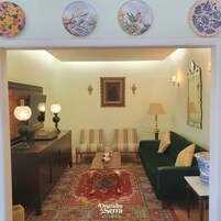 Uma sala de estar aconchegante com um sofá, cadeiras, uma mesa de centro e um tapete estampado. Há luminárias de parede, um quadro emoldurado e um espelho decorativo.