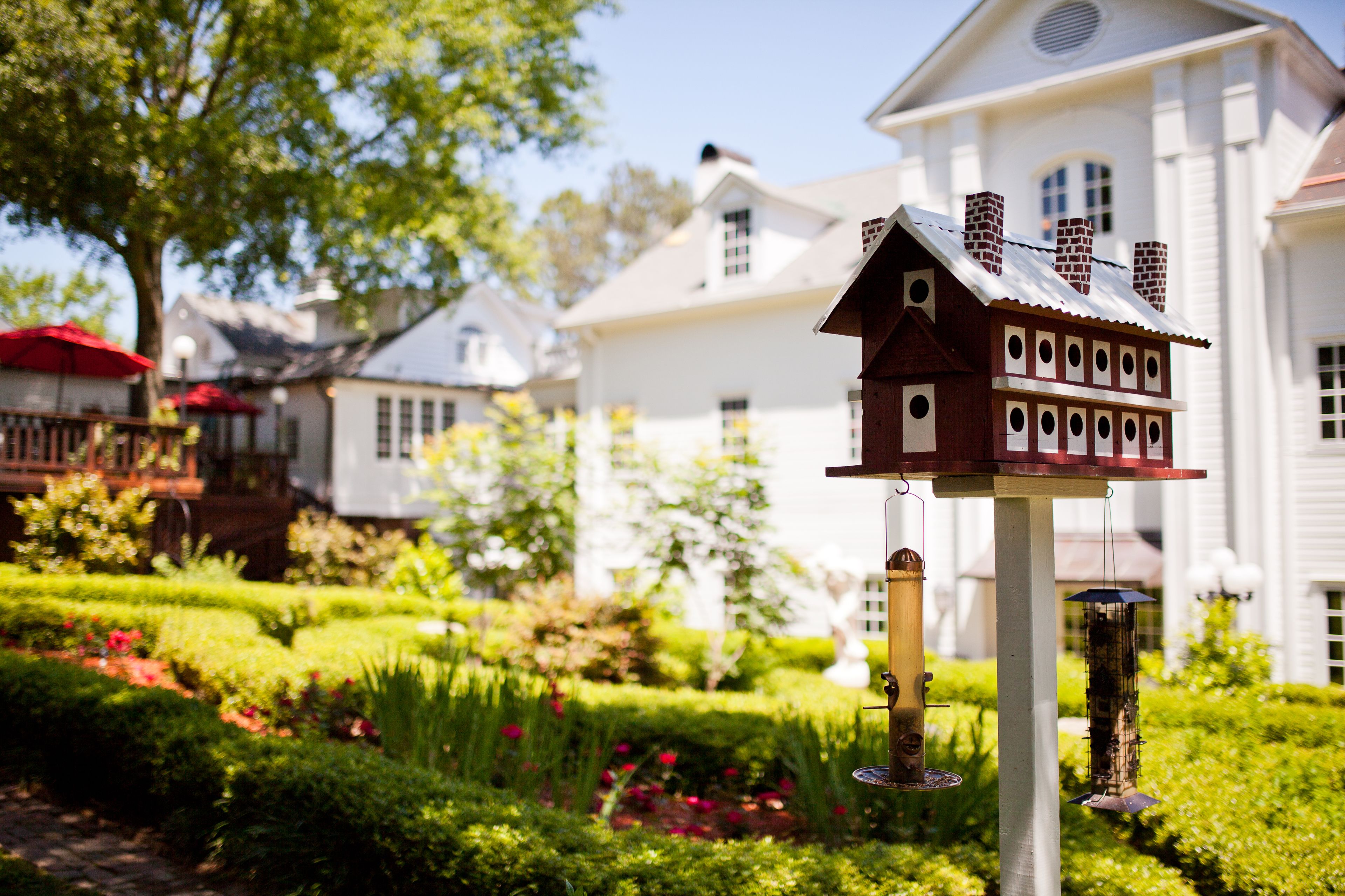 Um modelo de casinha de pássaro em um jardim, com uma casa branca ao fundo.