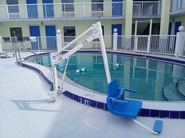 Uma área de piscina com elevador para piscina, uma cadeira de piscina azul e um edifício com varandas ao fundo.